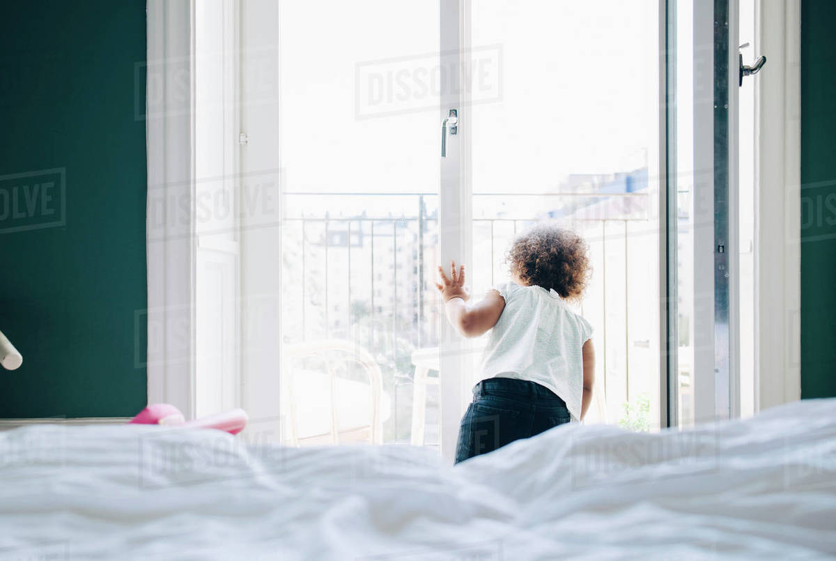 Cute girl looking through window while standing in bedroom at home ...