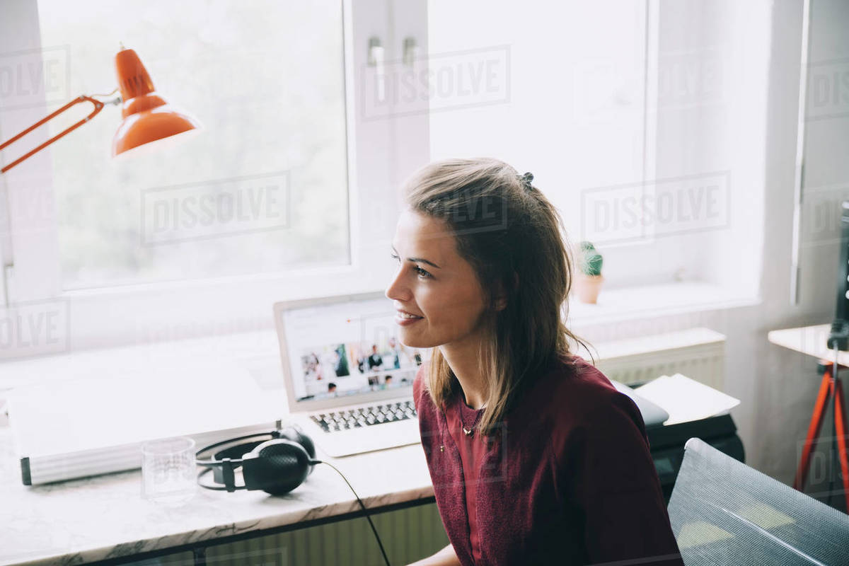 Confident businesswoman looking away while sitting at desk in office ...