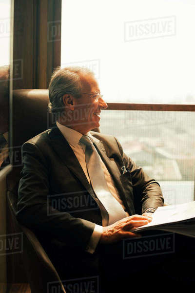 Smiling mature lawyer with documents looking through window at office ...