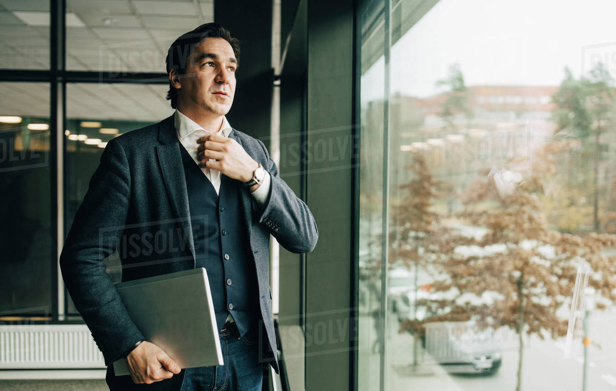 Businessman adjusting collar while standing with laptop by window in office - Royalty-free Stock ...