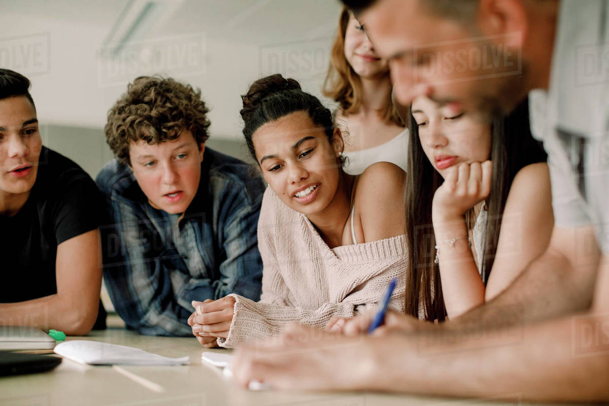 Teacher writing in book while teenage students leaning over table in ...