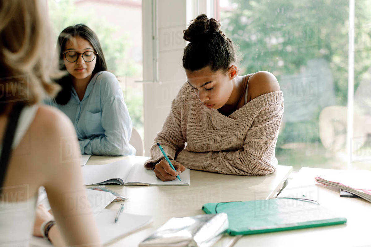Teenage student writing while sitting by table against glass window in ...