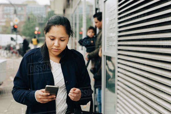 Mid adult woman using mobile phone while walking on sidewalk in city ...