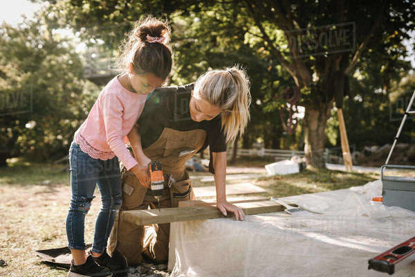 Daughter using drill machine on wooden plank by mother in backyard ...