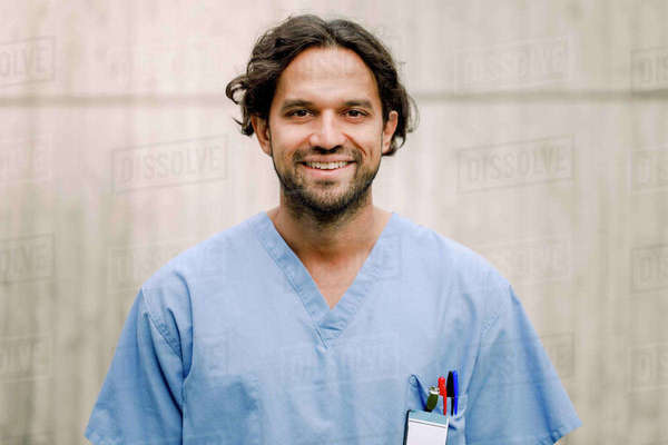 Portrait of smiling male nurse standing against wall in hospital ...