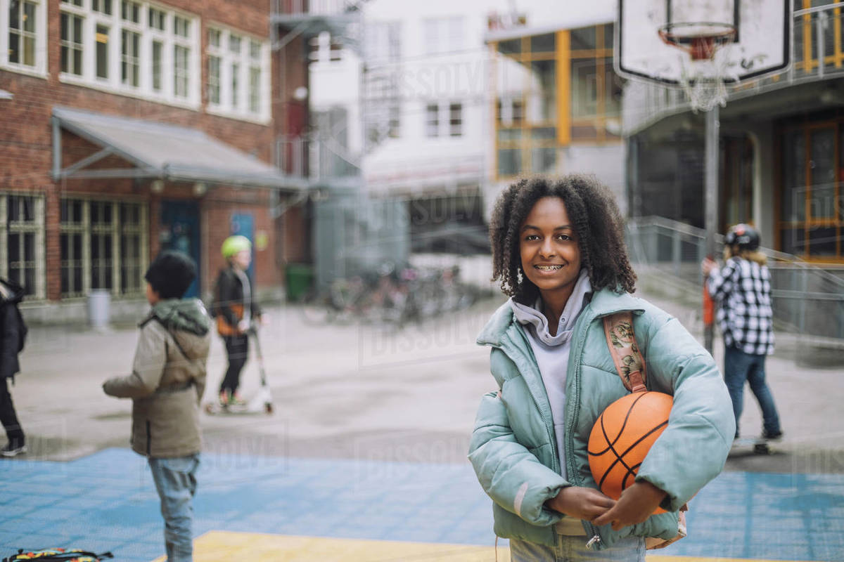 Portrait of smiling girl with basketball standing in sports court at ...