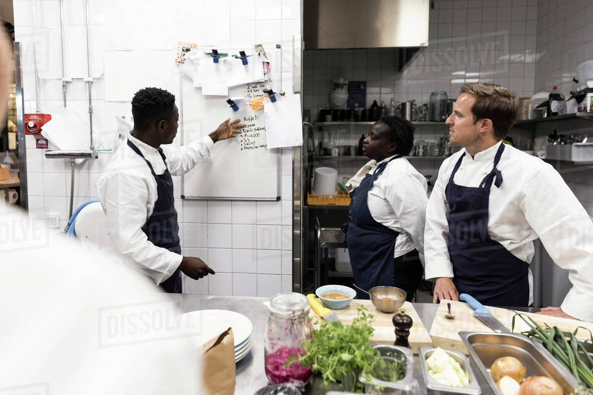 Male chef explaining to colleagues over whiteboard in kitchen of ...