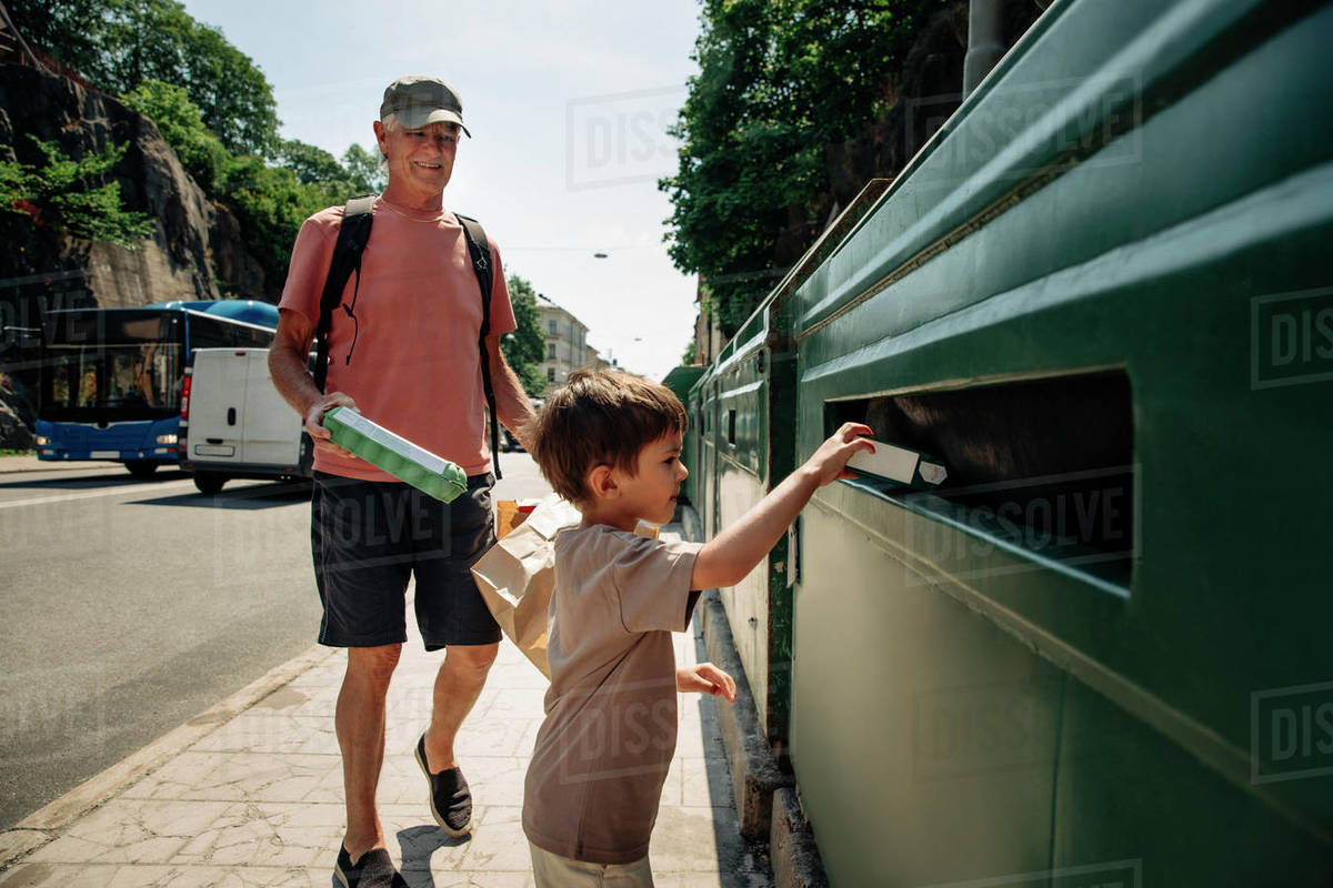 Boy trashing garbage in bin with grandfather at sidewalk - Royalty-free ...