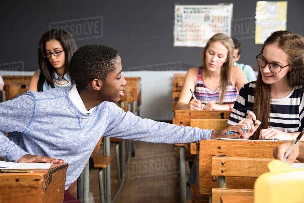 Two Students Sitting At Desk In Classroom Helping Each Other With Work Two Students Sitting At Desk In Classroom Helping Each Other With Work