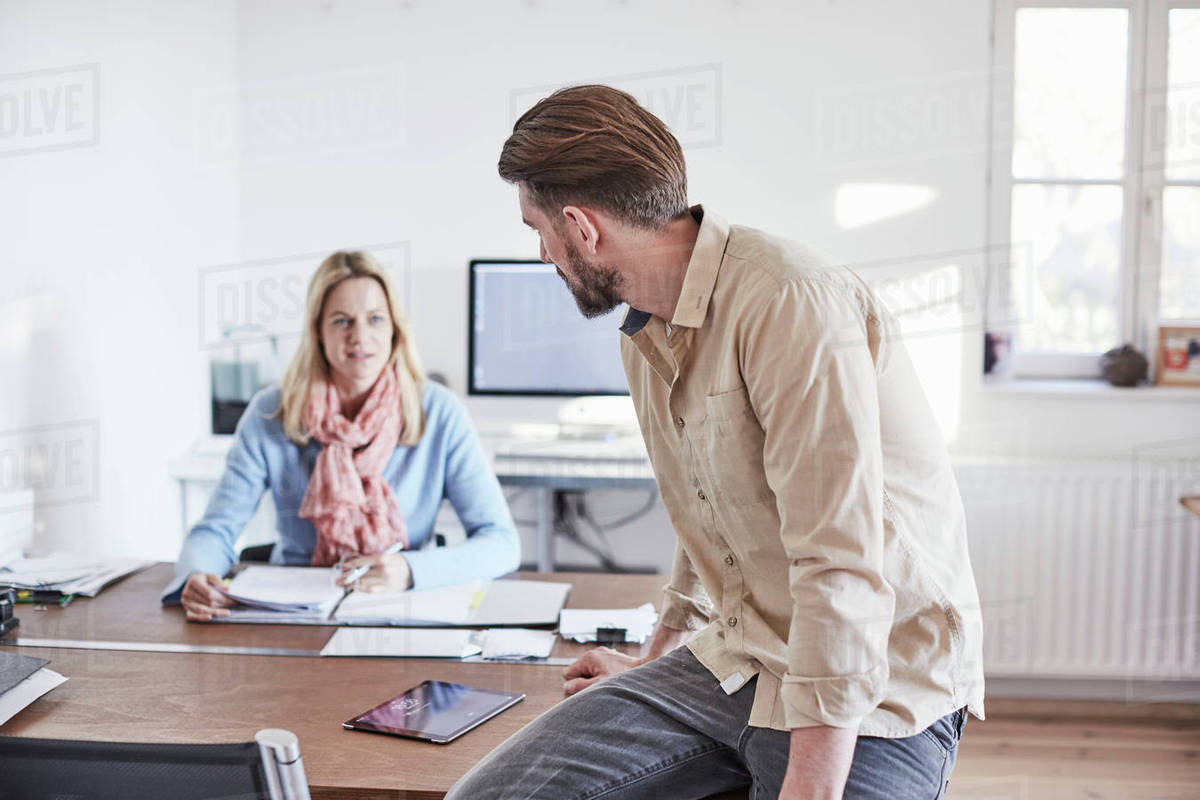 Man sitting on desk in office talking to colleague - Royalty-free Stock ...