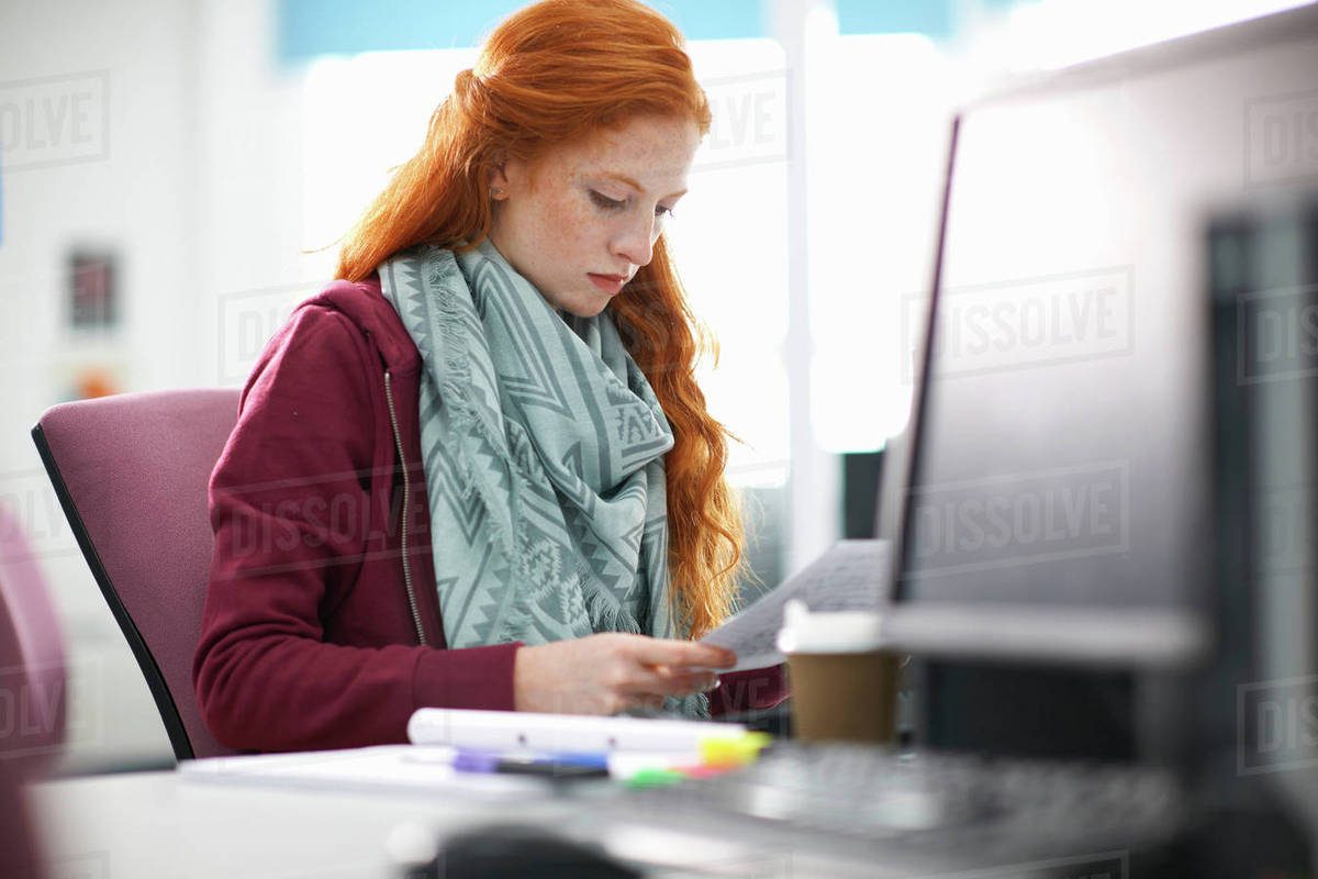 Young female college student at computer desk reading paperwork - Stock ...