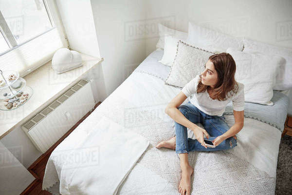 High level view of beautiful young woman sitting on bed gazing through ...