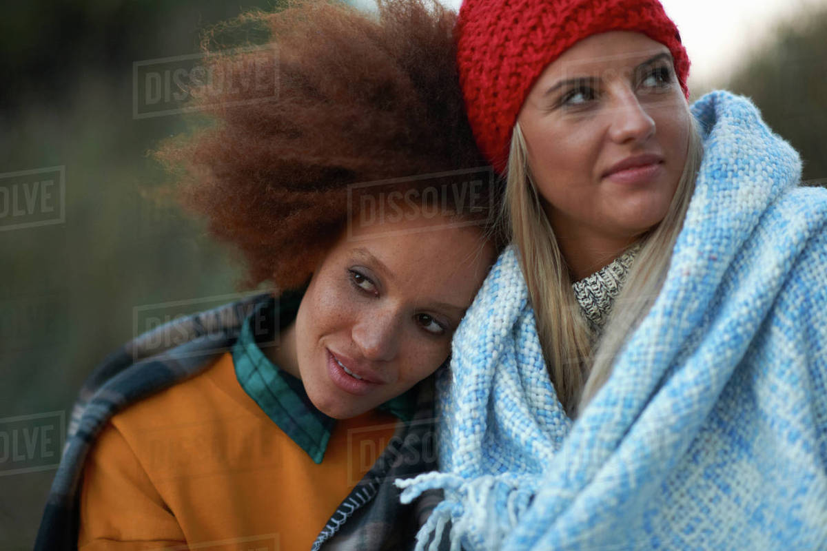 Portrait of two young women huddled in blanket on beach at dusk ...