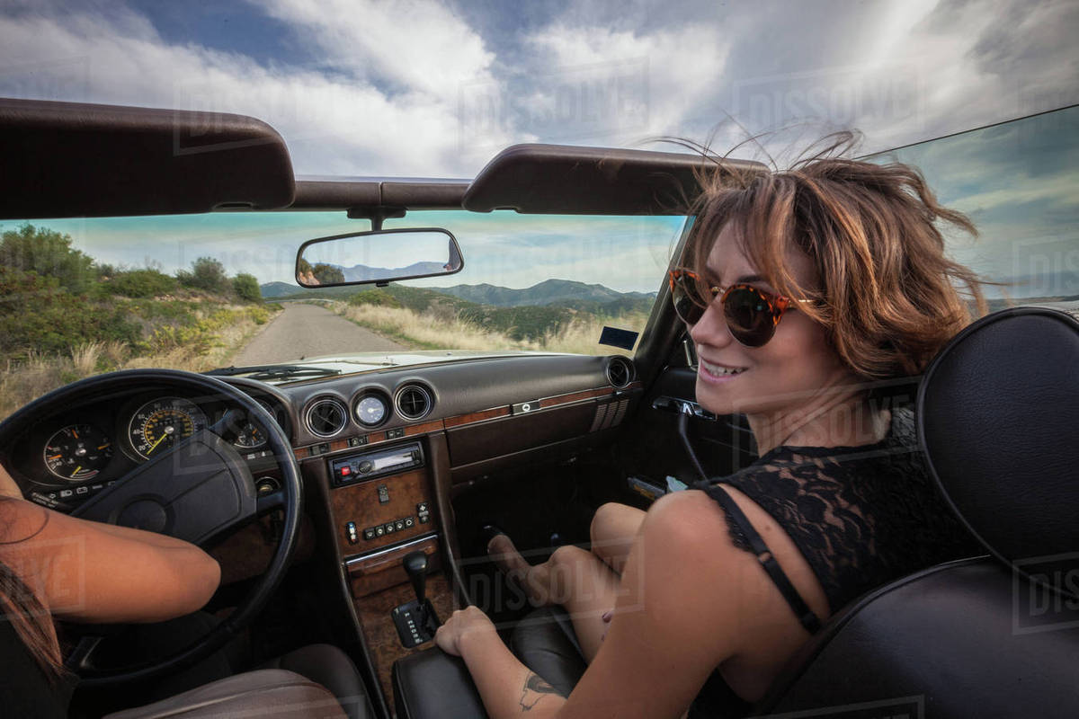 Two young women in convertible car, driving along scenic road, rear ...