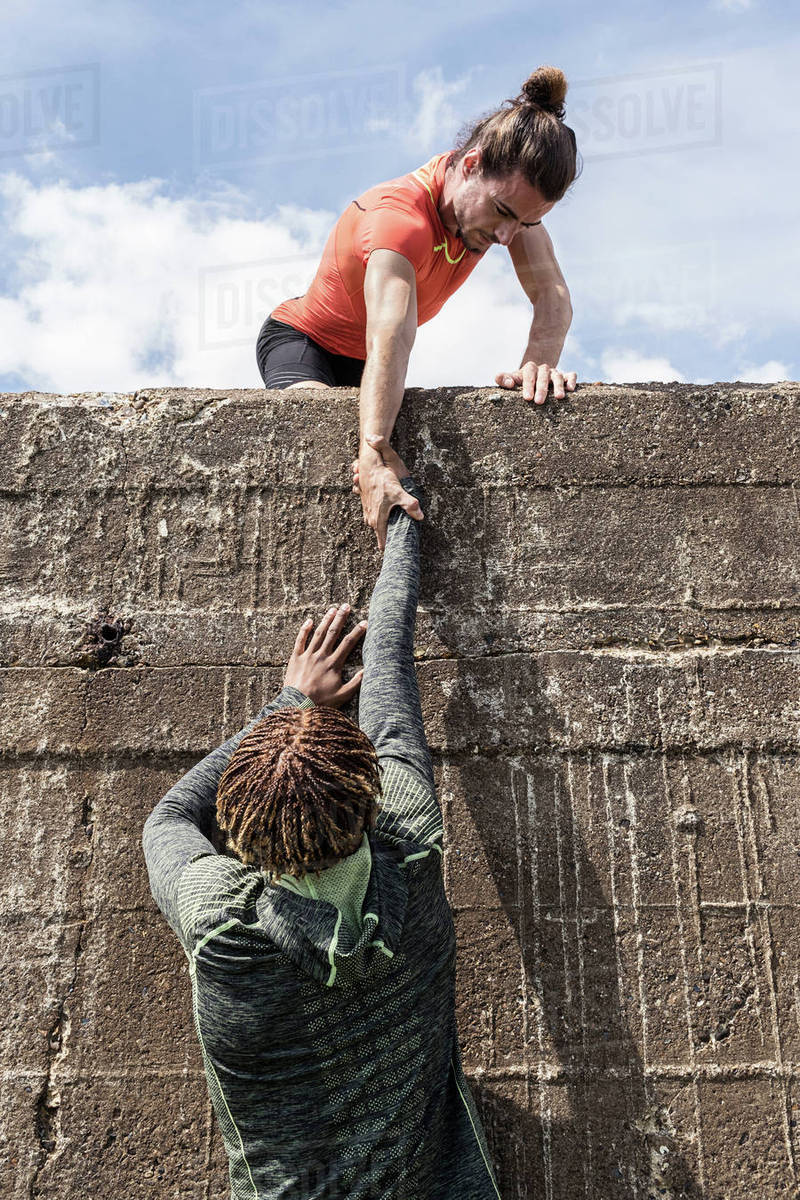 Young male free climber at top of sea wall helping friend climb up ...