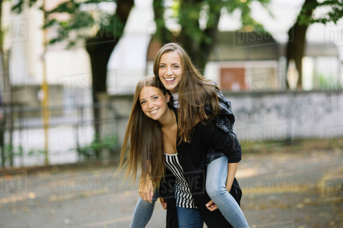 Portrait of young woman giving best friend a piggy back in park ...