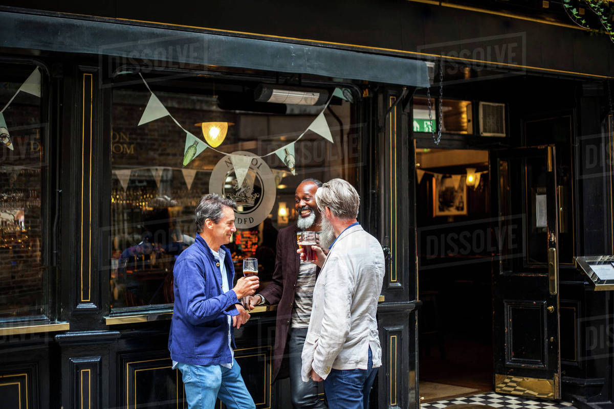 Three mature men, standing outside pub, holding beer glasses, laughing ...