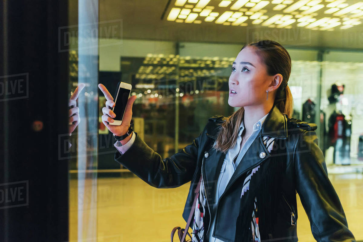 Woman looking in store window, holding smartphone - Stock Photo - Dissolve