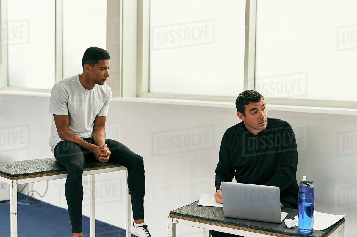 Athlete relaxing on table top listening to coach - Stock Photo - Dissolve