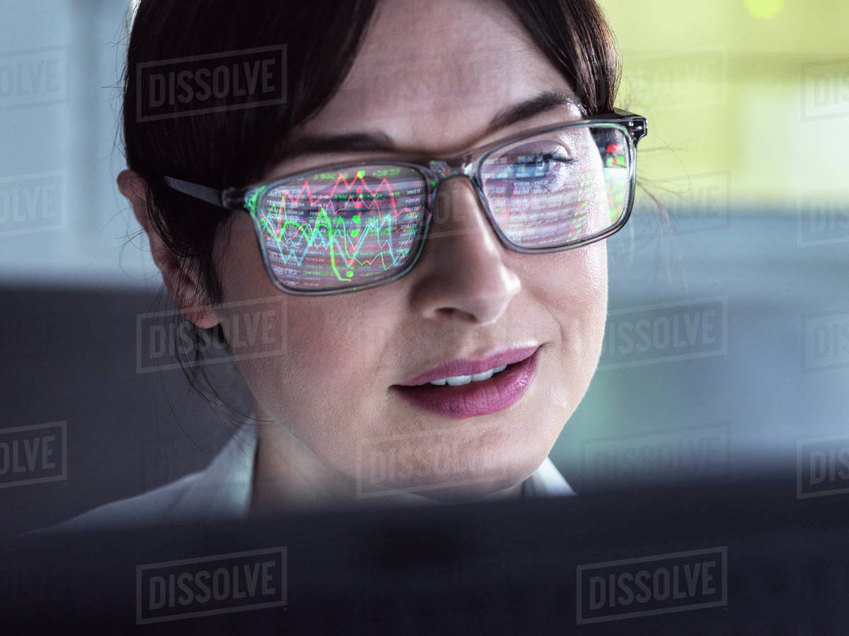 A woman looks closely at a monitor, her spectacles reflecting graphs ...