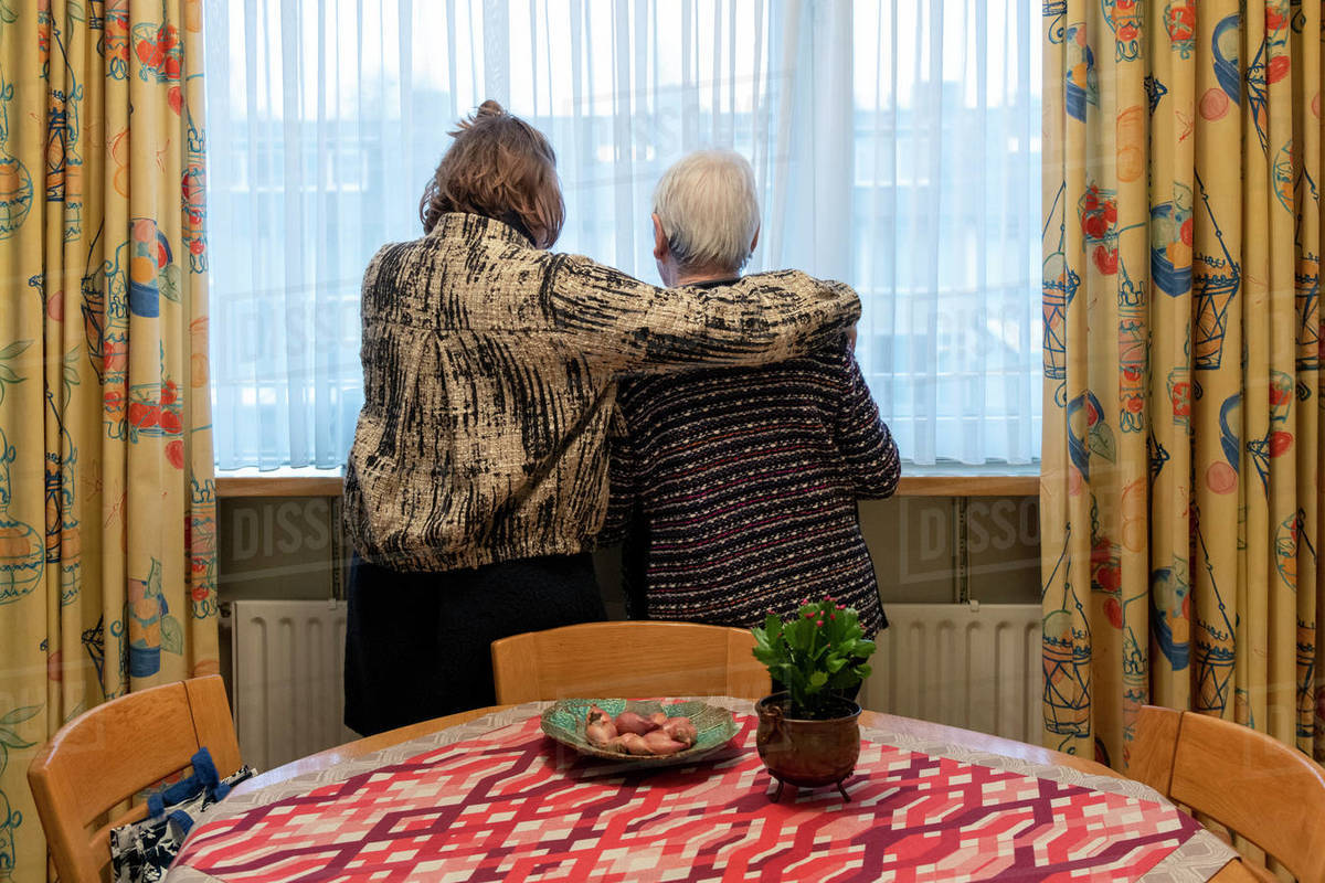 Two elderly women embrace while looking out the window, a cozy room ...