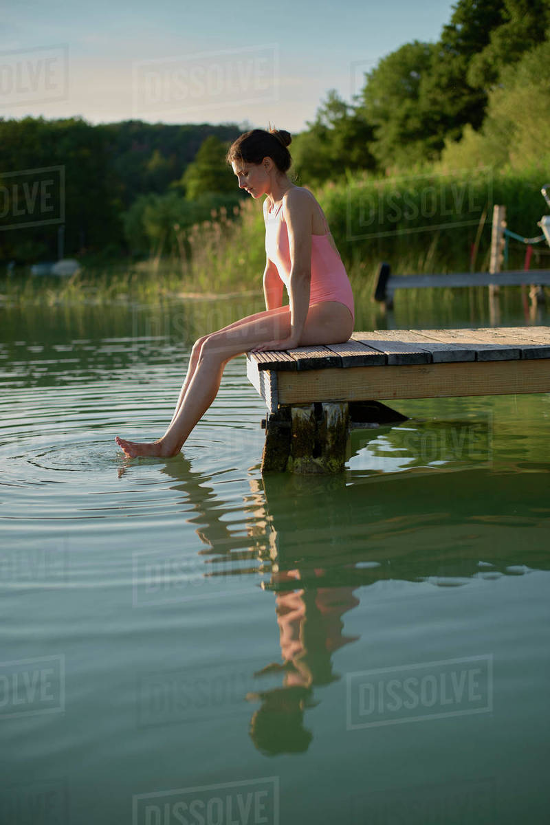 Woman in a pink swimsuit sitting on a wooden dock, dipping her feet in ...