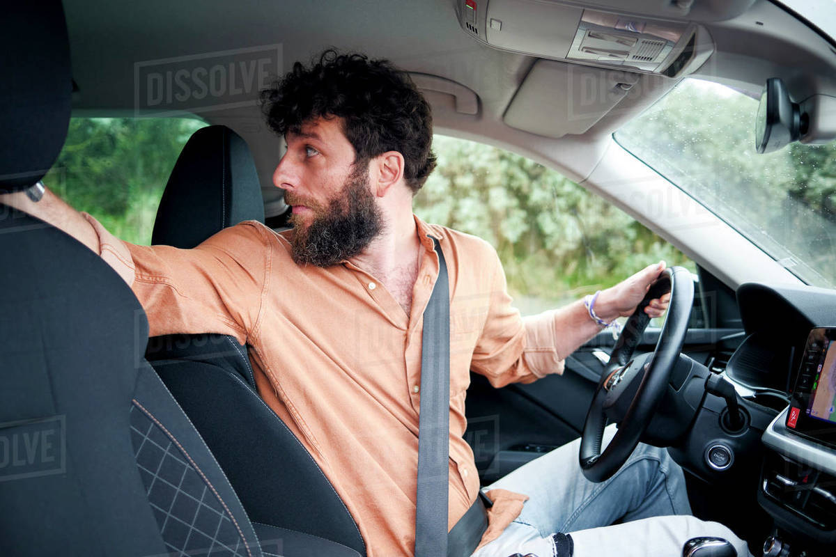 A bearded man driving a car looks back with a puzzled expression ...