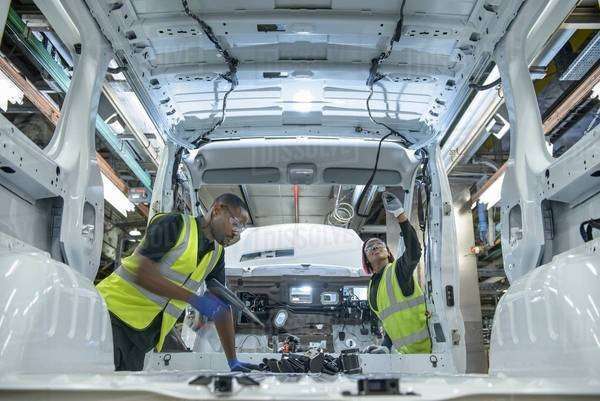 Car workers wearing high visibility jackets fitting parts on vehicle on ...