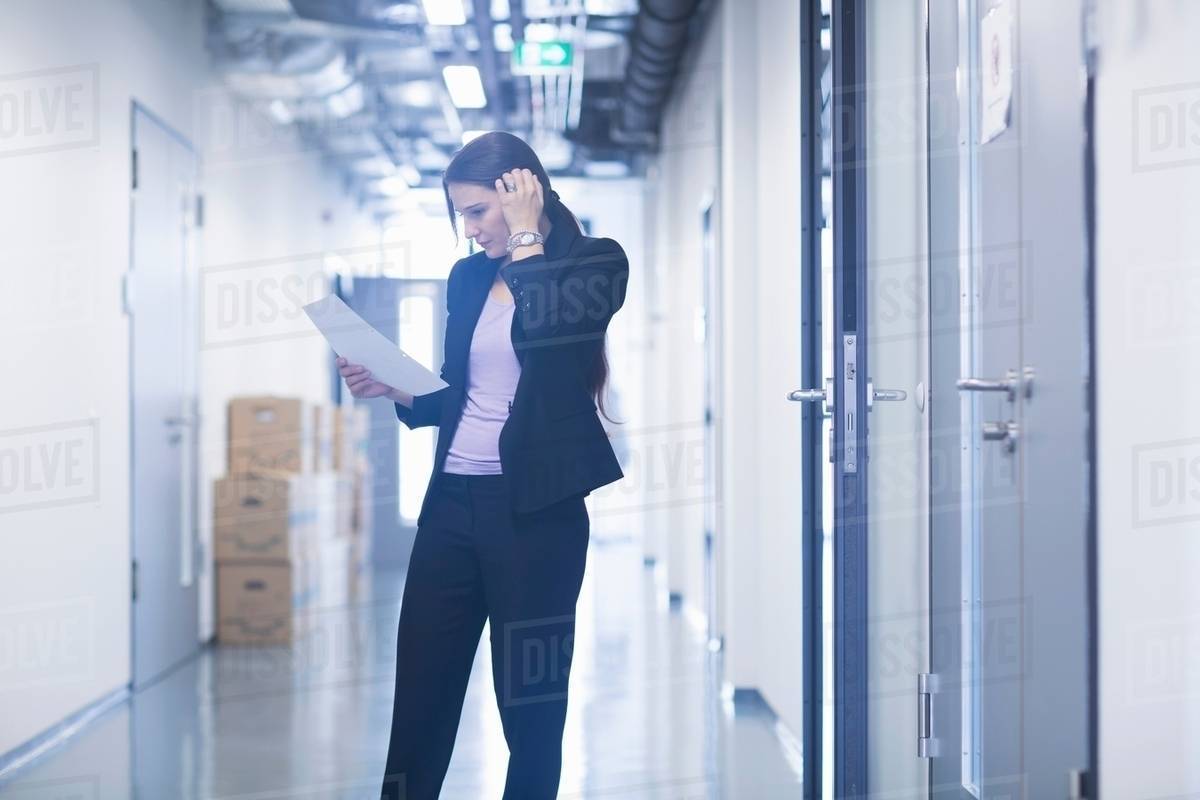 Young woman standing in office corridor looking down at paperwork ...