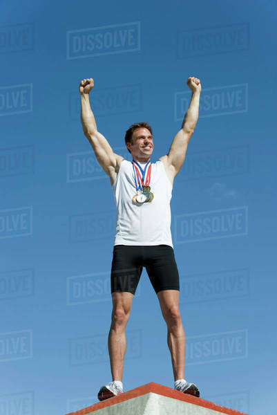 Male athlete standing on winner's podium with arms raised in victory ...