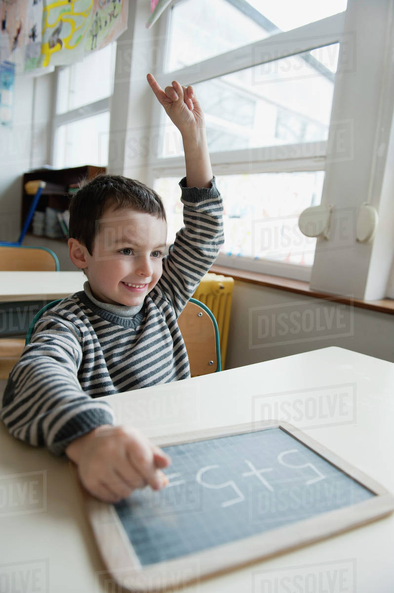 Boy sitting at desk with chalkboard, hand raised - Royalty-free Stock ...