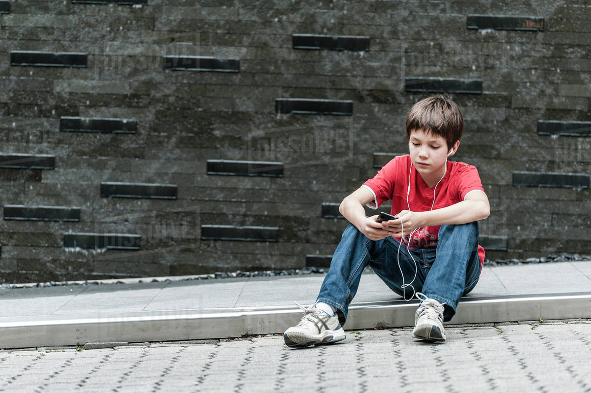 Boy sitting on sidewalk listening to music with earphones - Royalty ...