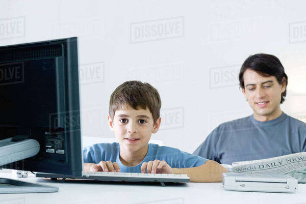 Little boy using computer, smiling at camera, father reading newspaper ...