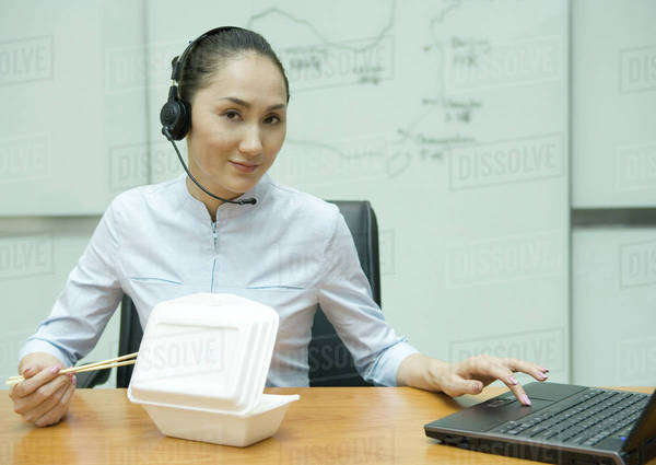Businesswoman eating takeout food, wearing headset, and using laptop ...