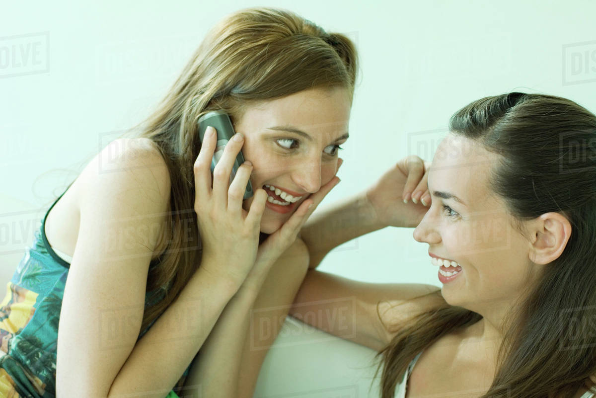 Two young friends smiling at each other, one using cell phone - Royalty ...