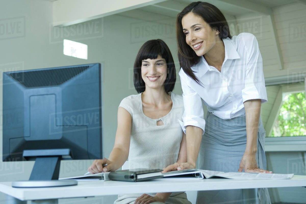 Two businesswomen looking at computer monitor together, smiling ...