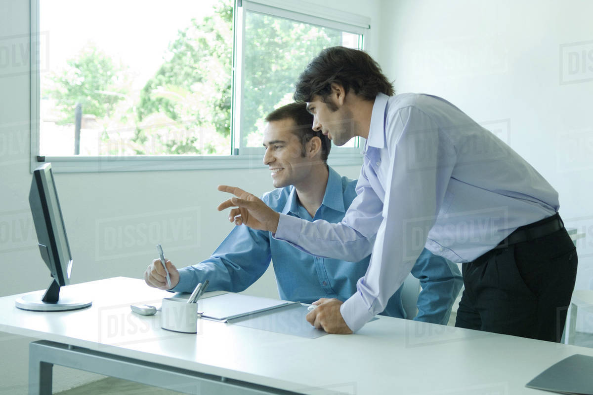 Businessman sitting at desk facing monitor while colleague stands next ...