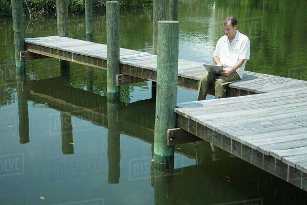 Man sitting on dock, using laptop computer, looking down, smiling ...