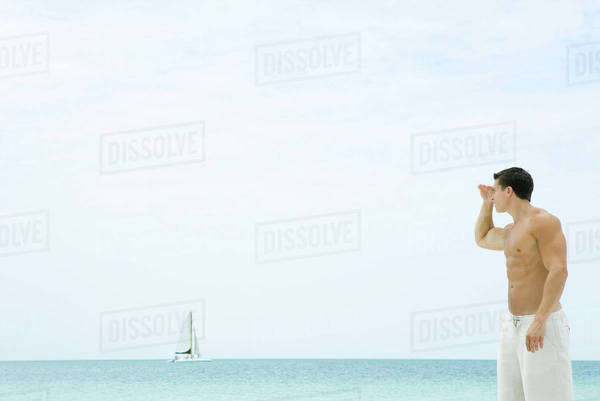 Man standing at the beach, looking at view, sailboat in the distance ...