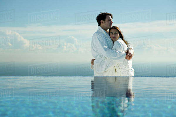 Couple embracing at edge of infinity pool, both wearing bathrobes ...