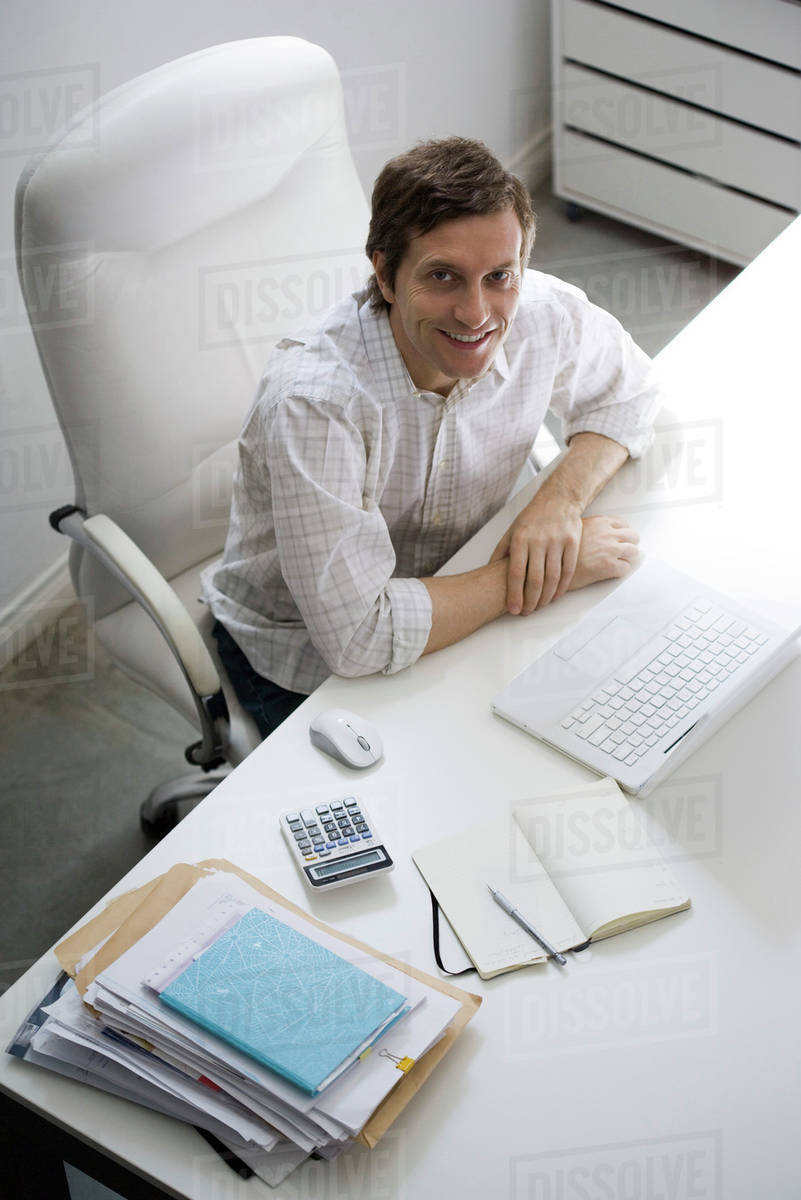 Man sitting at desk, smiling up at camera - Stock Photo - Dissolve
