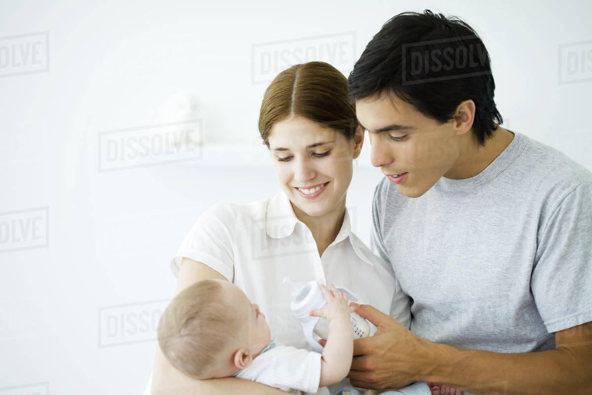 Young parents smiling down at baby, father helping baby hold bottle ...
