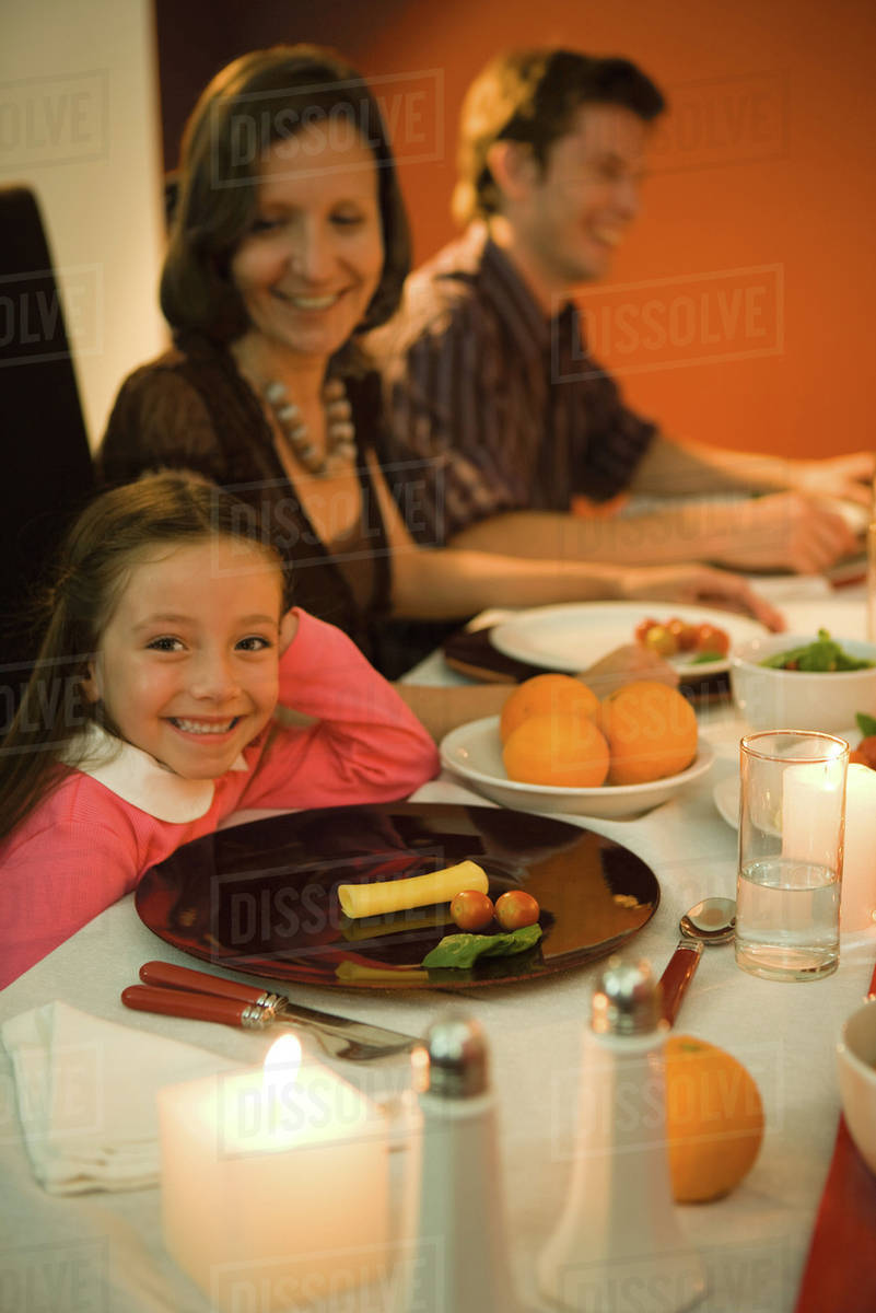 Girl sitting at dinner table, smiling at camera, adults in background ...