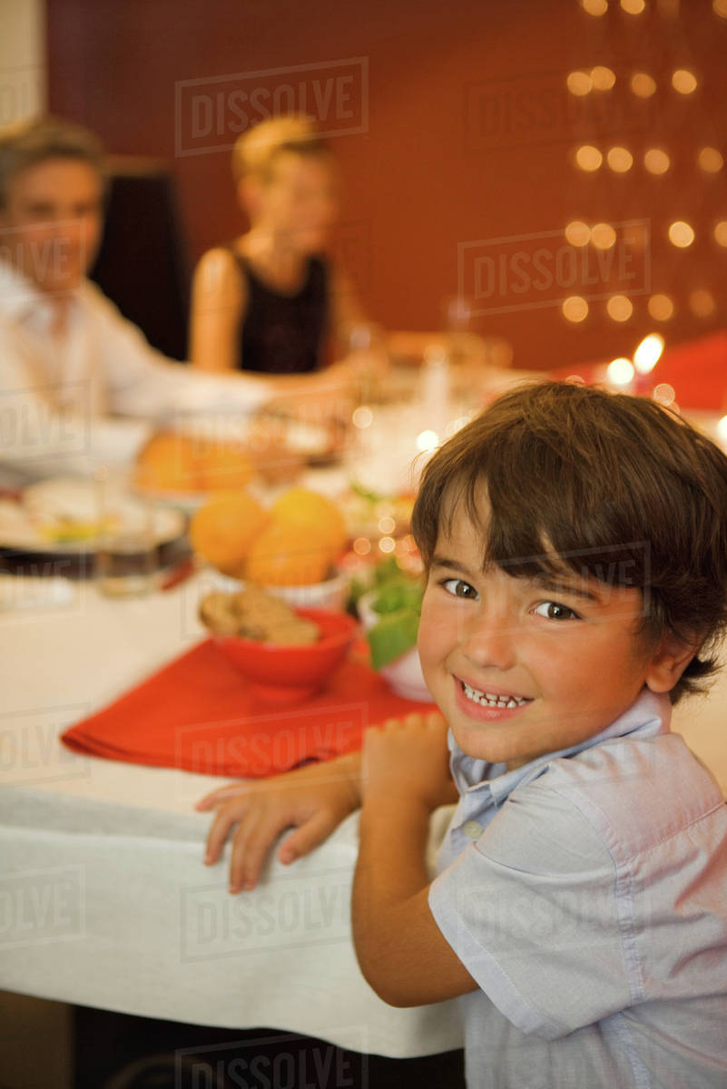Boy sitting at dinner table, smiling at camera, adults in background ...