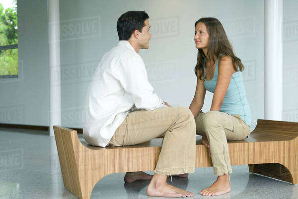 Young couple sitting face to face on bench, smiling at each other ...