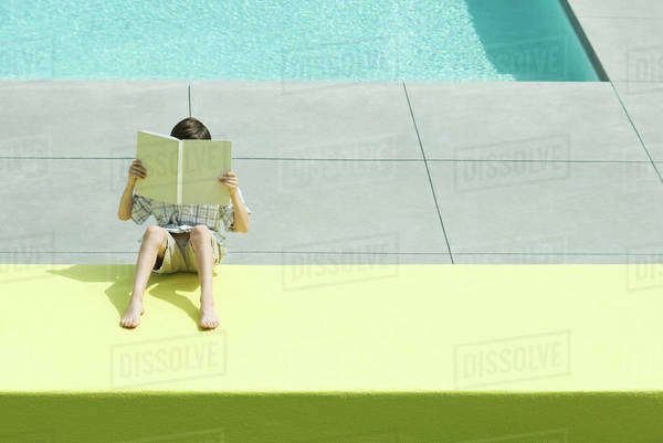 Boy reading beside swimming pool, face obscured by book, high angle ...