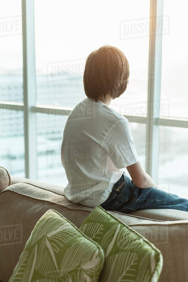 Teenage boy sitting on edge of sofa, looking out window, rear view ...