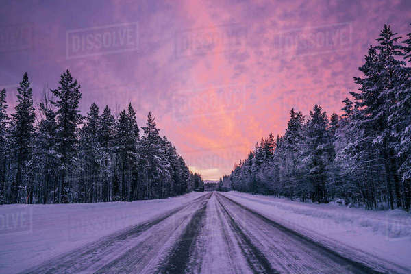 Remote winter road through snow covered forest trees against dramatic ...