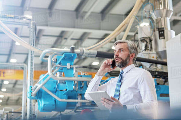 Male supervisor with digital tablet talking on cell phone in factory ...