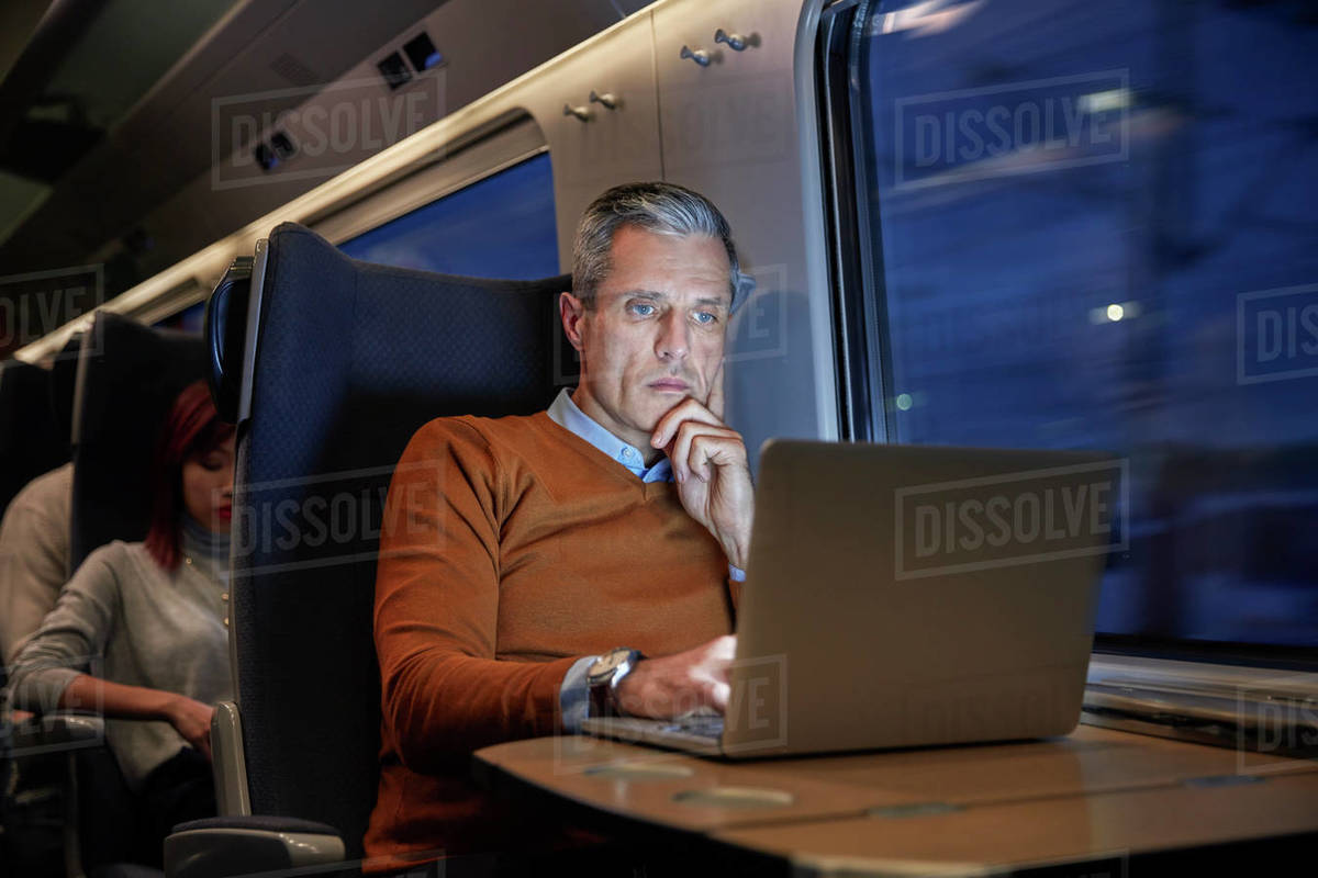 Focused businessman working at laptop on passenger train at night ...